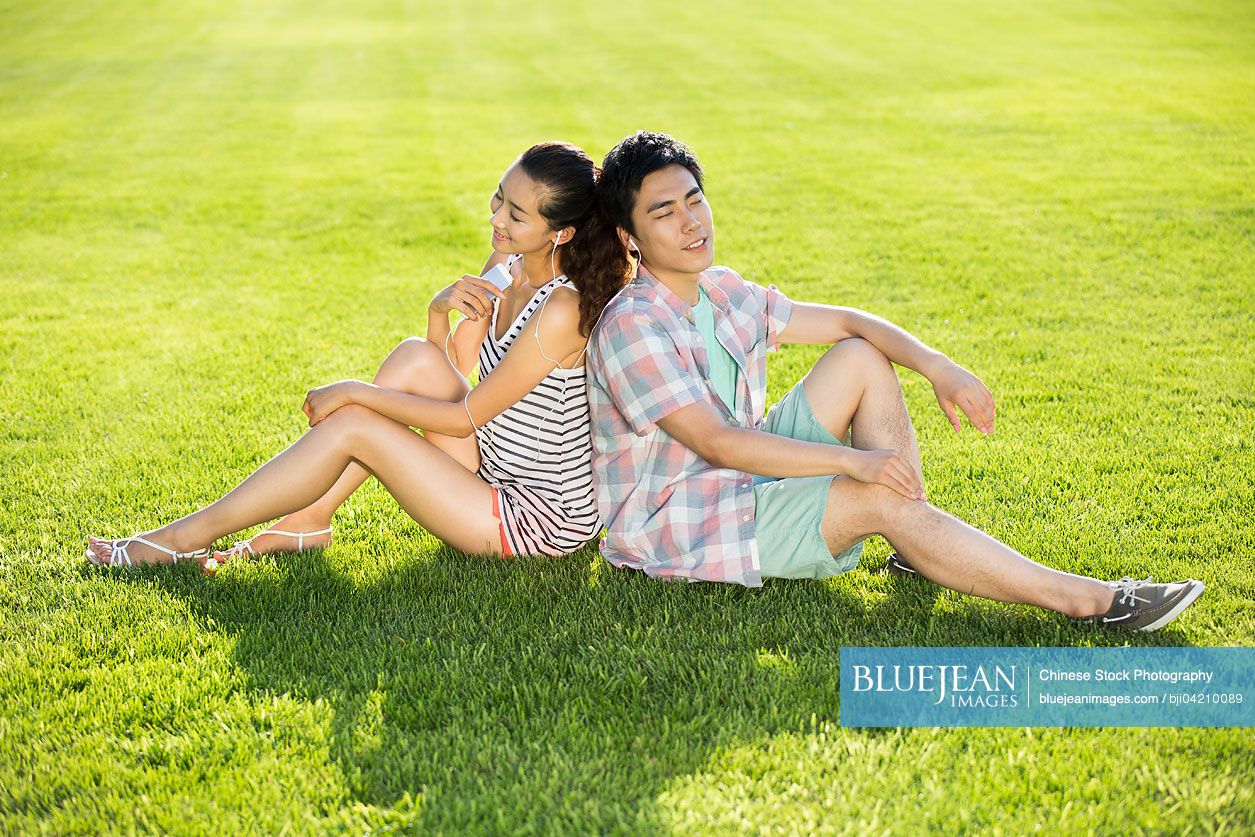 Cheerful young Chinese couple sitting back to back on grass listening ...