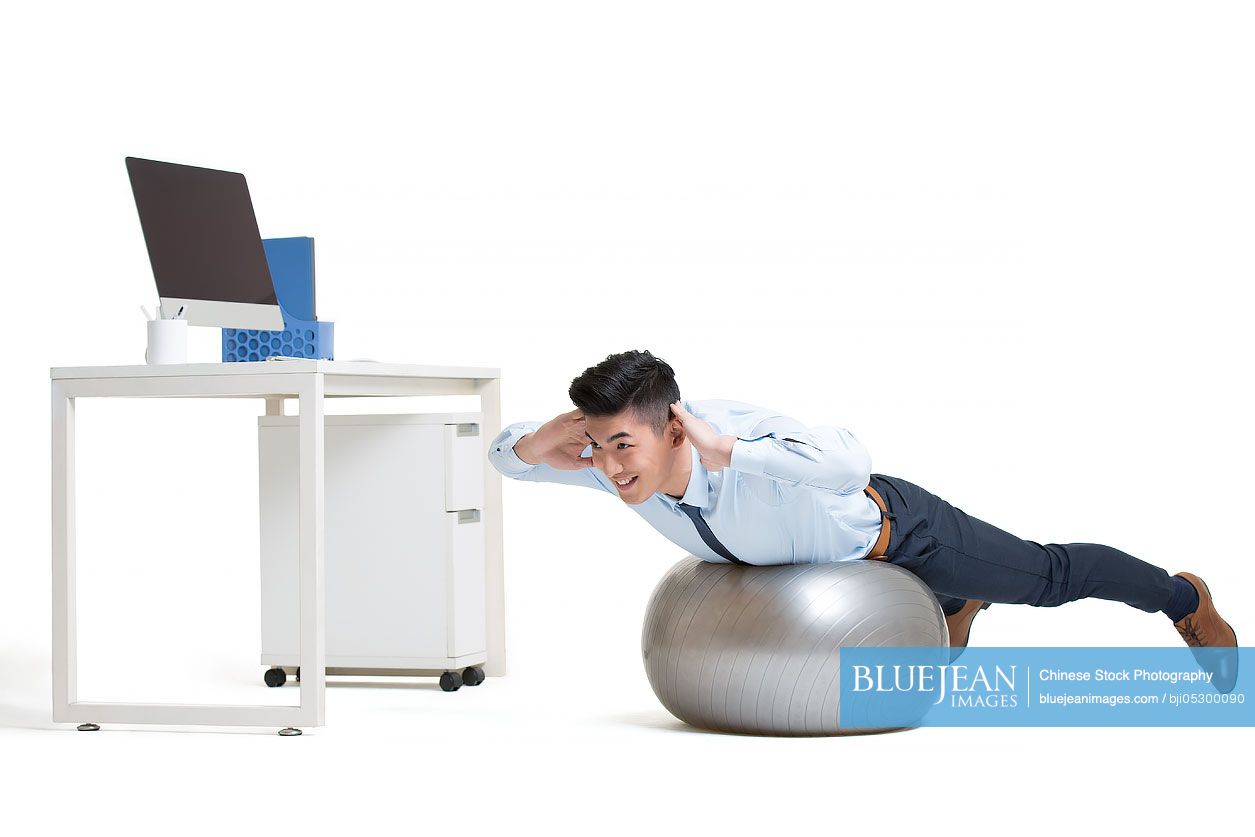 Young Chinese businessman exercising in office while sitting on fitness ...