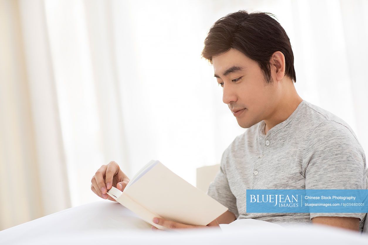 Happy young Chinese man reading book in bedroom-High-res stock photo ...