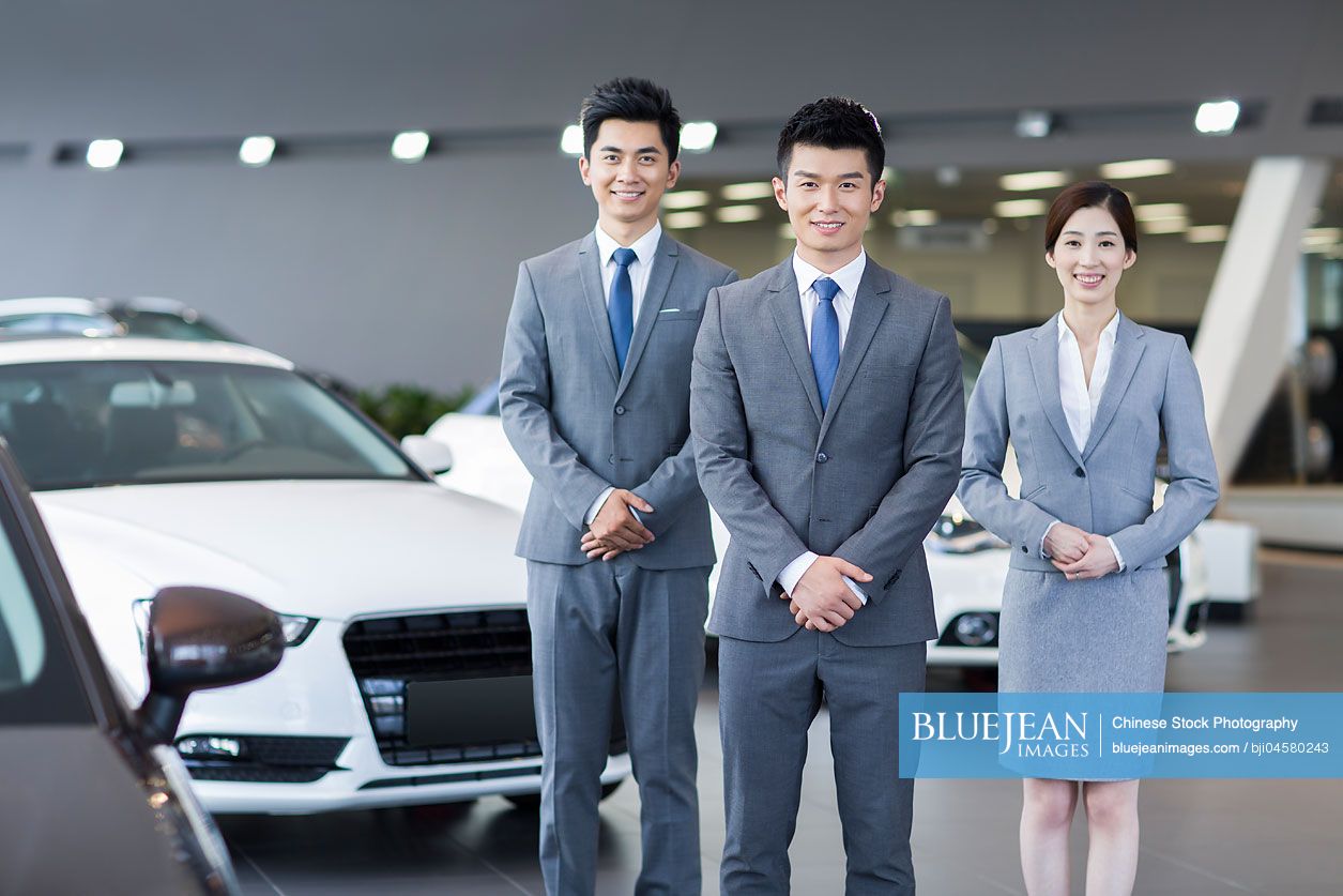 Confident Chinese salespeople standing with new cars in showroom-High ...