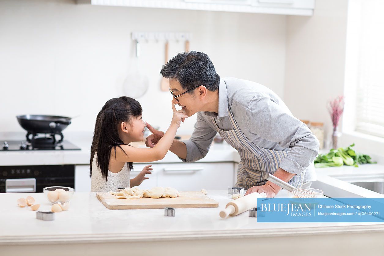 Happy little Chinese girl and grandfather baking cookies in kitchen ...