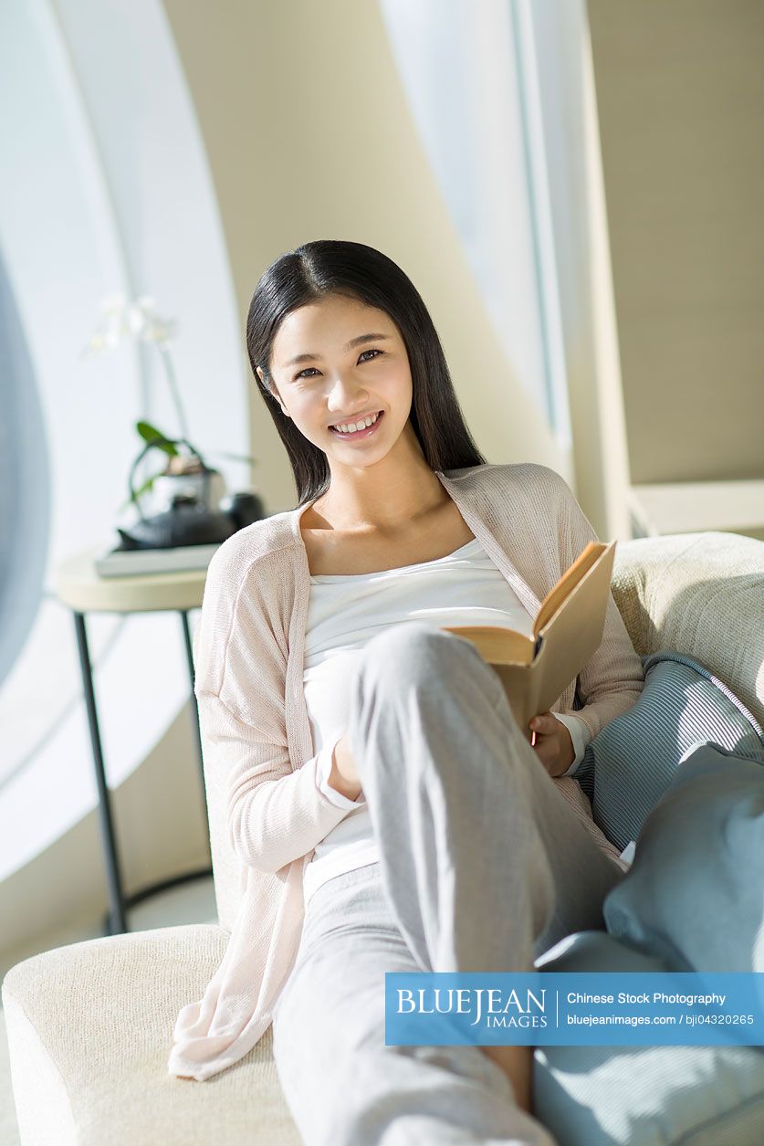 Happy young Chinese woman sitting on sofa with a book-High-res stock ...
