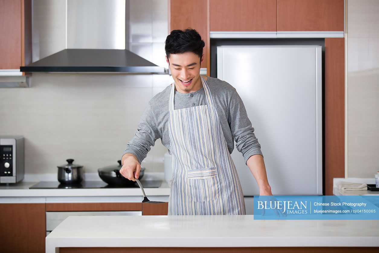 Cheerful Chinese young man cooking in kitchen-High-res stock photo for download